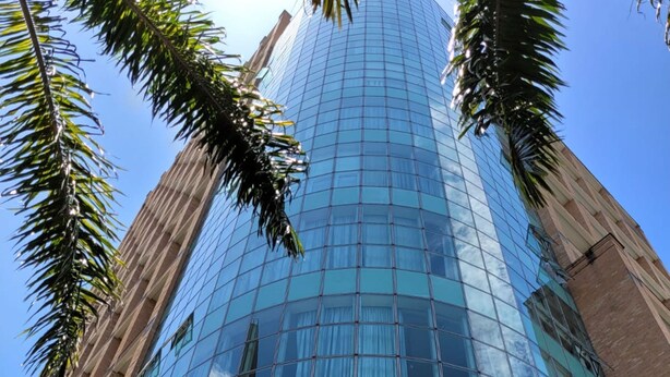 Um edifício alto e moderno com fachada de vidro refletiva, rodeado por palmeiras.