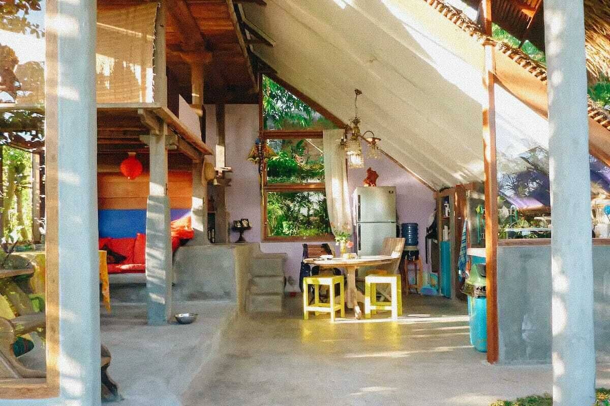 Uma área externa coberta com mesa, cadeiras, geladeira e janela com vista para o verde.