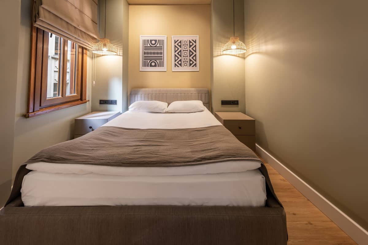 Quarto com cama, mesas de cabeceira, janela com persianas e quadros nas paredes.