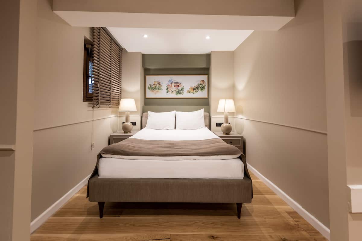 Quarto de hotel estreito com cama, abajures, janela com persianas e uma obra de arte na parede.
