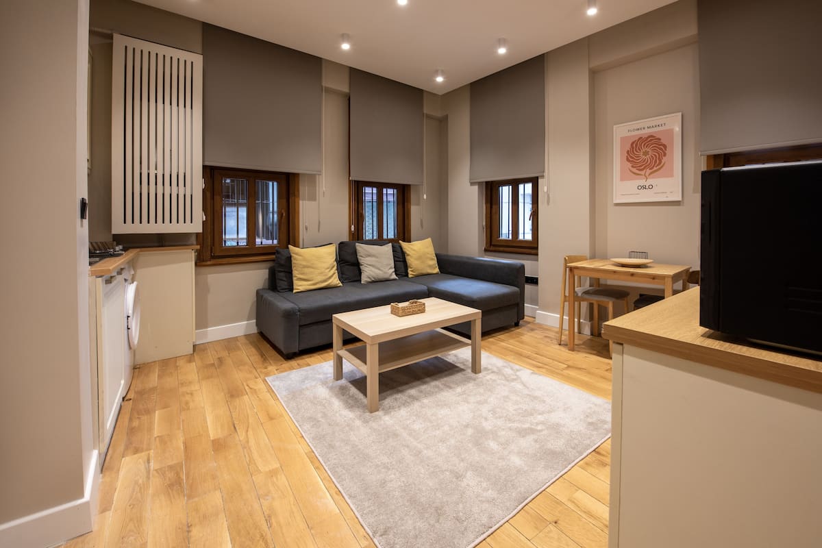 Uma sala moderna com um sofá cinzento, uma mesa de centro de madeira e uma área de cozinha com uma TV.