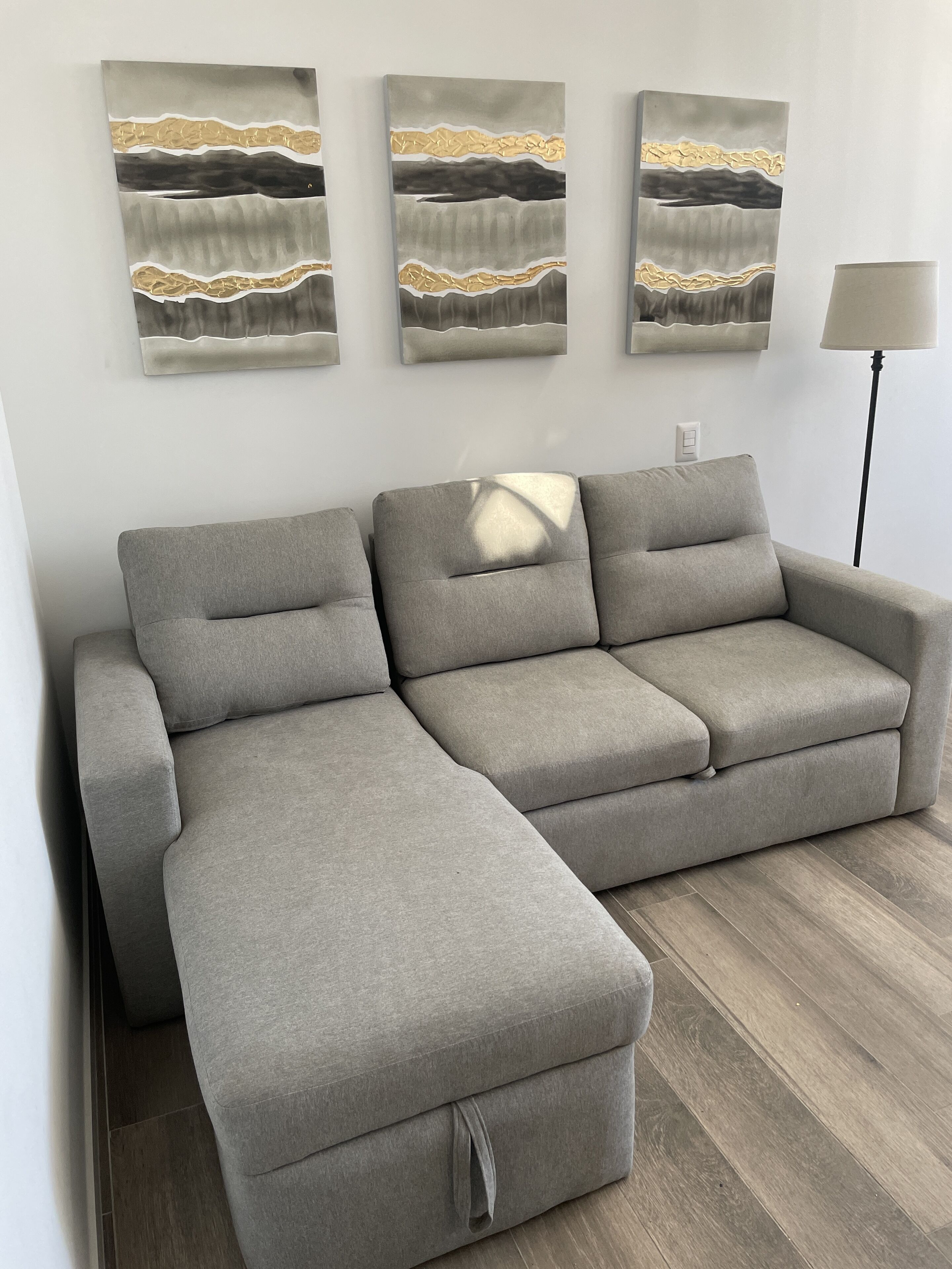 Um sofá modular cinza com apoio para os pés, uma luminária de chão e três obras de arte abstratas na parede.