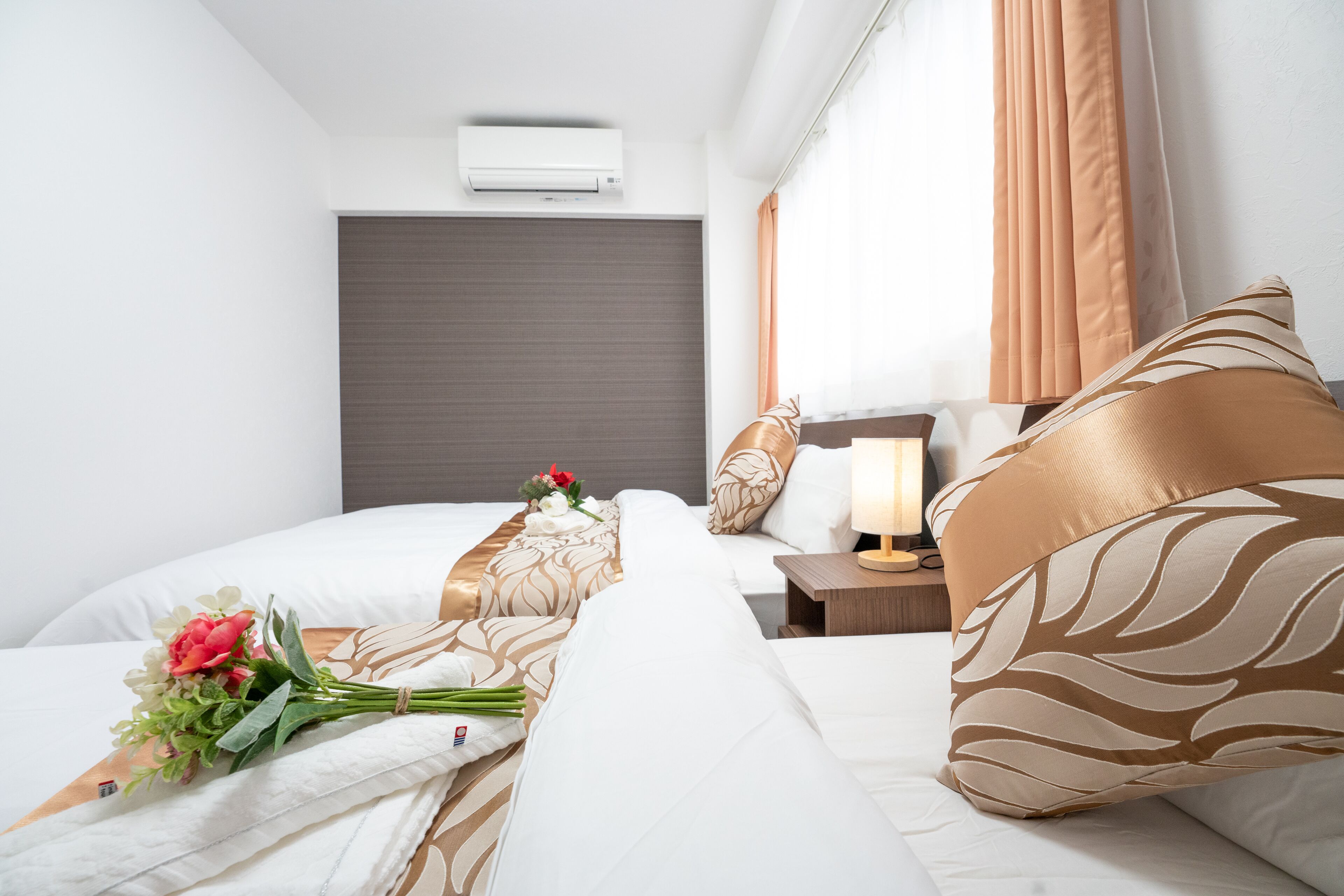 Quarto de hotel com duas camas, uma mesa de cabeceira com abajur, um buquê de flores e uma janela com cortinas.