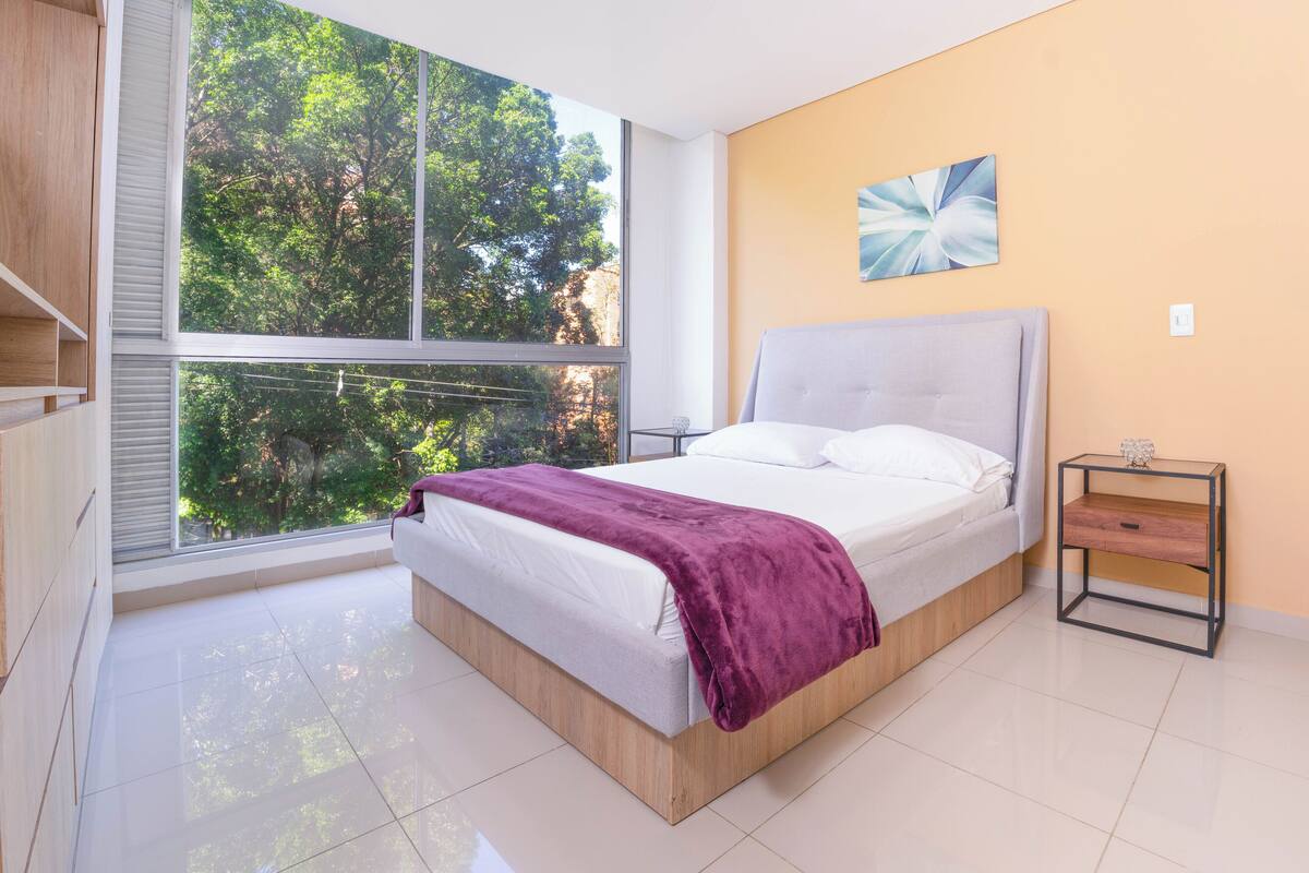 Quarto moderno com uma janela grande, uma cama com cabeceira branca, um cobertor roxo e uma pequena mesa de cabeceira com abajur.
