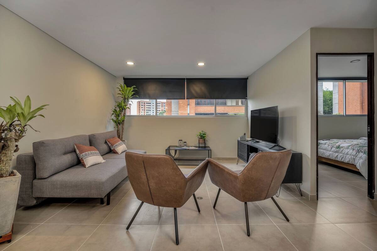 Uma sala de estar moderna com um sofá cinzento, duas cadeiras marrons, uma televisão e uma escrivaninha com uma planta.