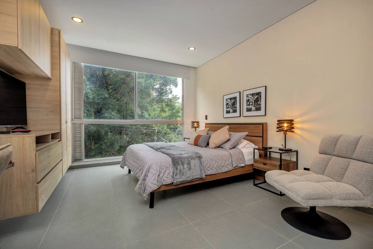 Um quarto moderno com uma janela grande, uma cama com colcha cinza, uma mesa de cabeceira de madeira, uma poltrona cinza e uma TV sobre um móvel de madeira.