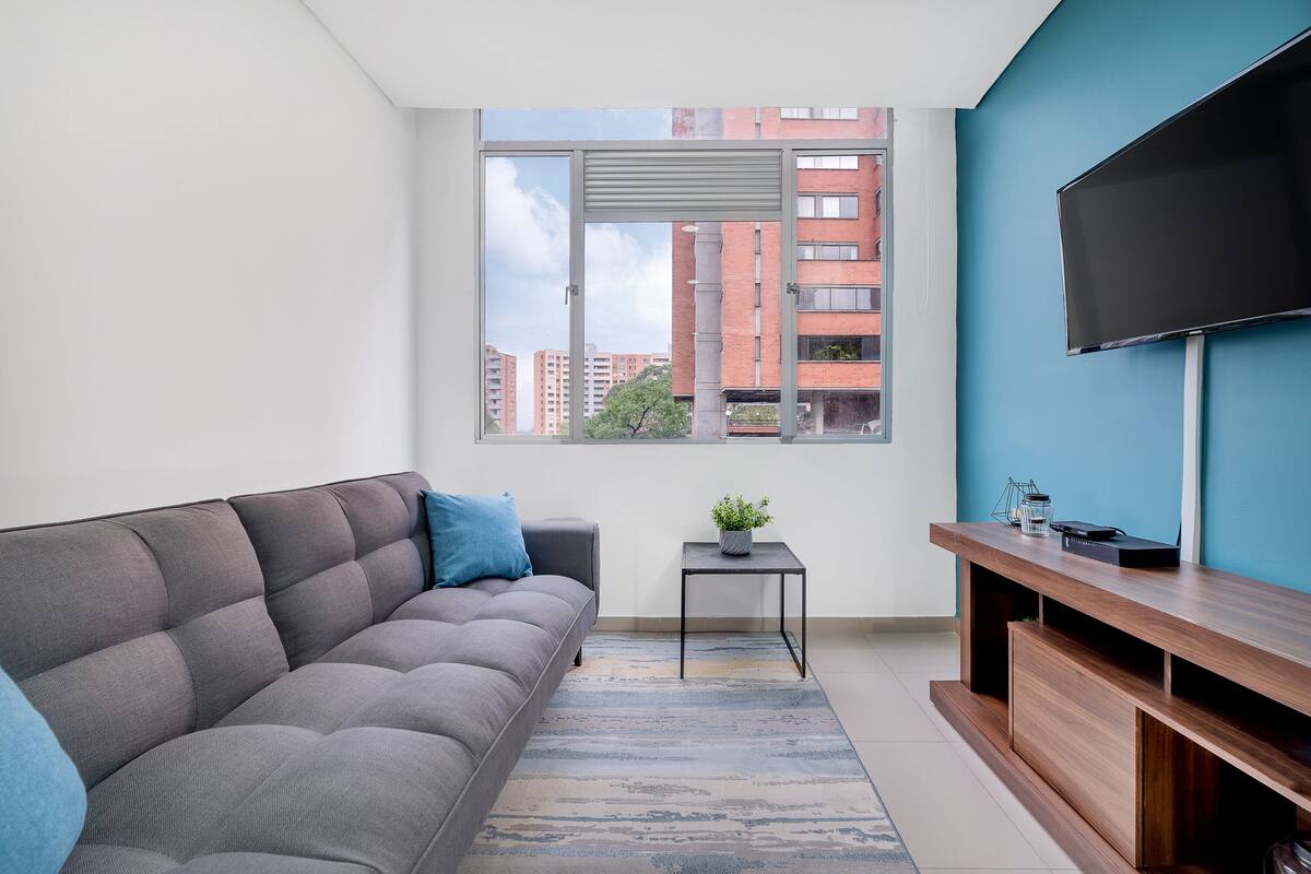 Uma sala de estar moderna com um sofá cinzento, uma mesinha de apoio com uma planta, um móvel de TV em madeira e uma janela grande com vista para edifícios.