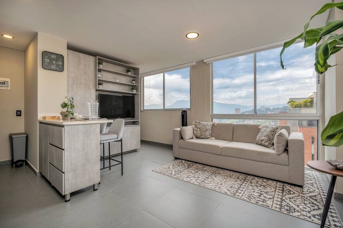 Um espaço de estar moderno com cozinha, sofá e uma grande janela com vista para a cidade.