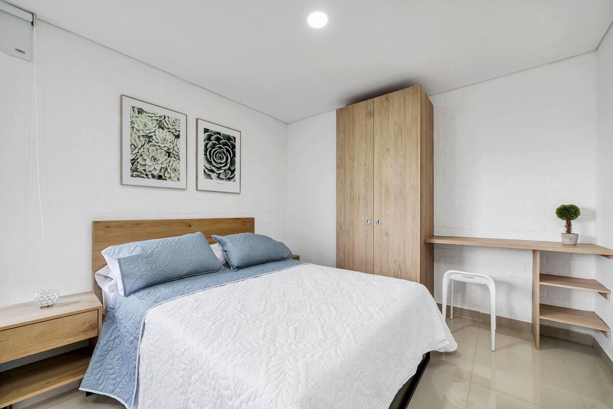 Quarto com cama, guarda-roupa de madeira, escrivaninha com cadeira e duas obras de arte penduradas na parede.