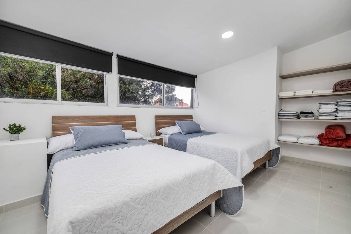 Quarto moderno com duas camas, janela com persiana, mesa de cabeceira com planta e prateleira com toalhas dobradas.