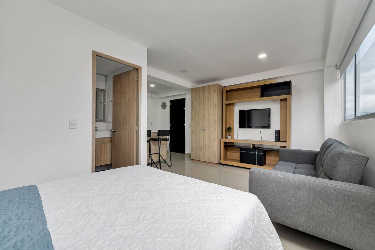 Quarto de hotel moderno com uma cama grande, um sofá cinzento, uma TV na parede e uma pequena área de kitchenette.