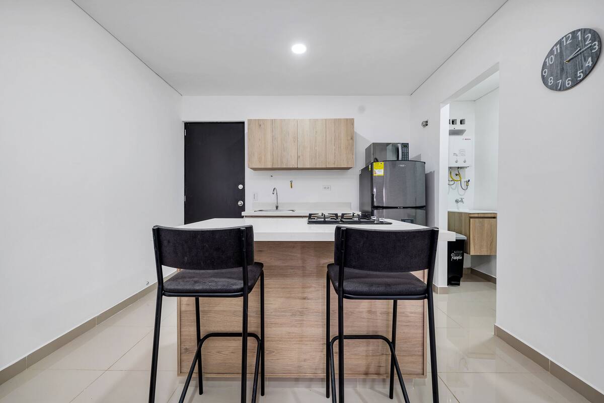 Uma cozinha moderna com dois bancos altos, um armário de madeira, uma geladeira de aço inoxidável e um relógio na parede.