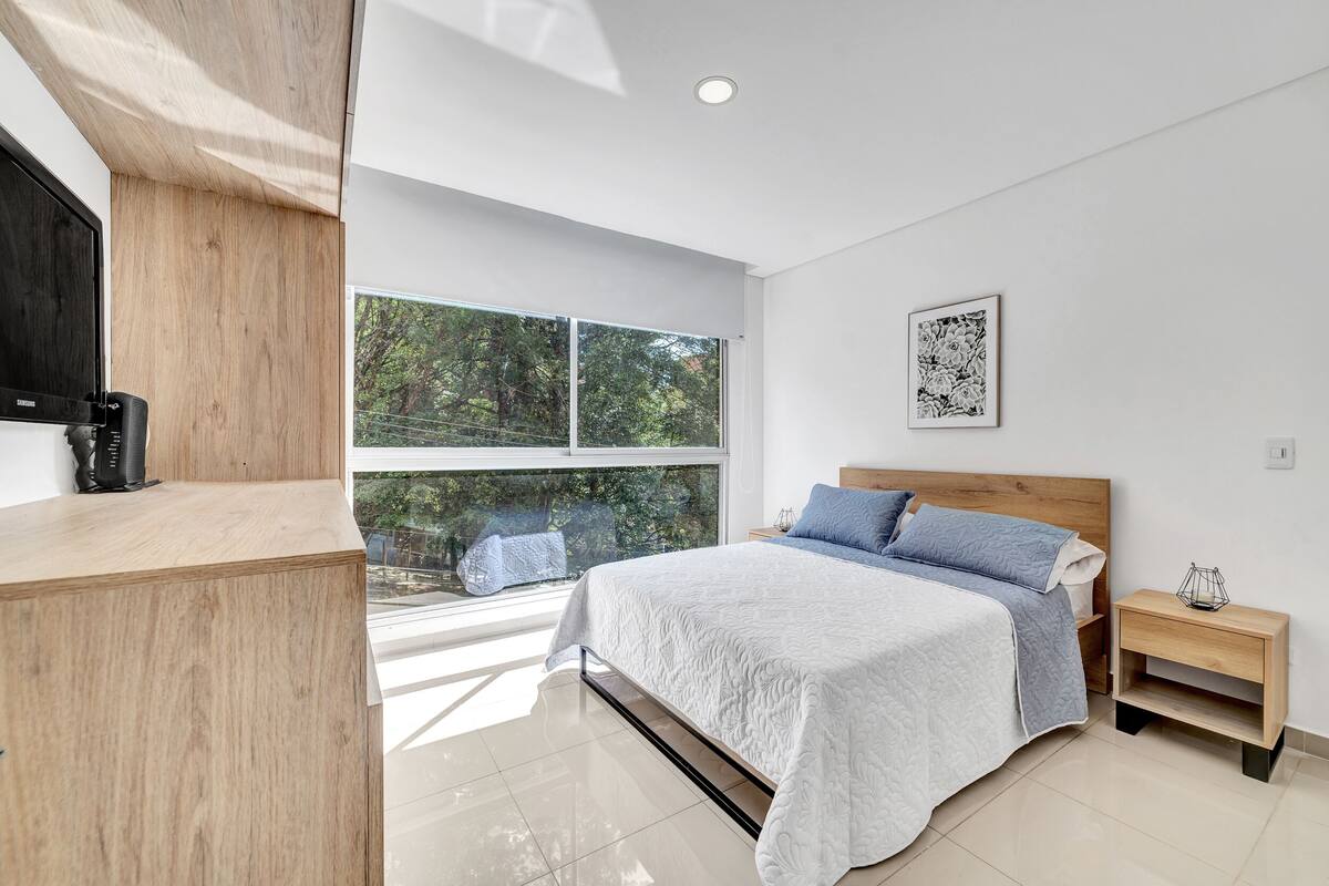 Um quarto moderno com uma cama grande, uma mesa de cabeceira de madeira, uma mesinha de apoio e uma janela com vista para árvores.