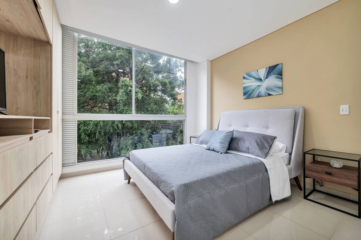 Quarto com uma janela grande, cama com roupa de cama cinzenta, mesa de cabeceira e guarda-roupa de madeira.