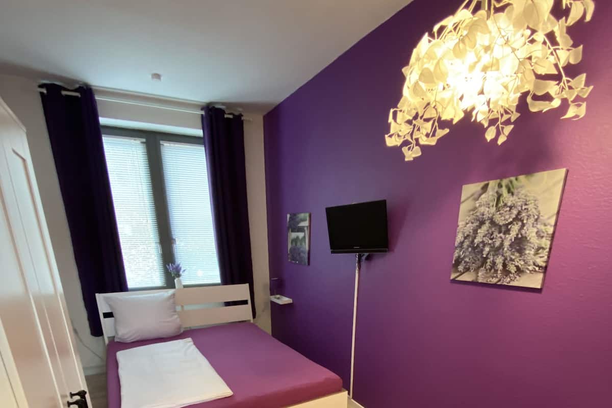 Quarto com cama, televisão, parede roxa e janela com cortinas.