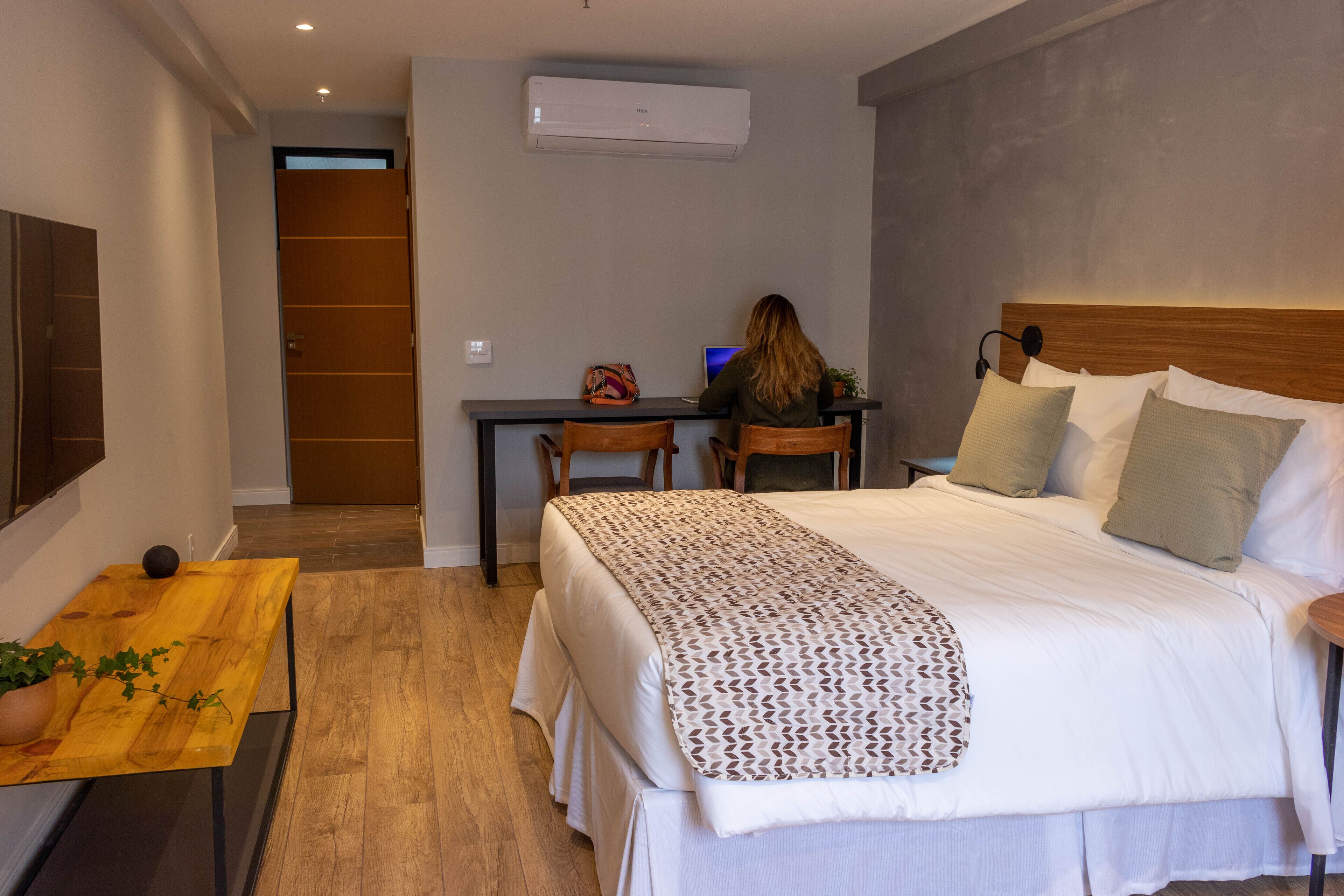 Quarto de hotel com cama, mesa de cabeceira, escrivaninha e cadeira.