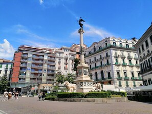 Uma praça pública com uma estátua de uma figura alada sobre uma coluna, rodeada por edifícios com varandas e persianas verdes.