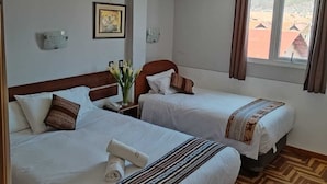 Quarto de hotel com duas camas, piso de madeira, janela com cortinas, quadro emoldurado na parede e uma pequena mesa com um vaso de flores.