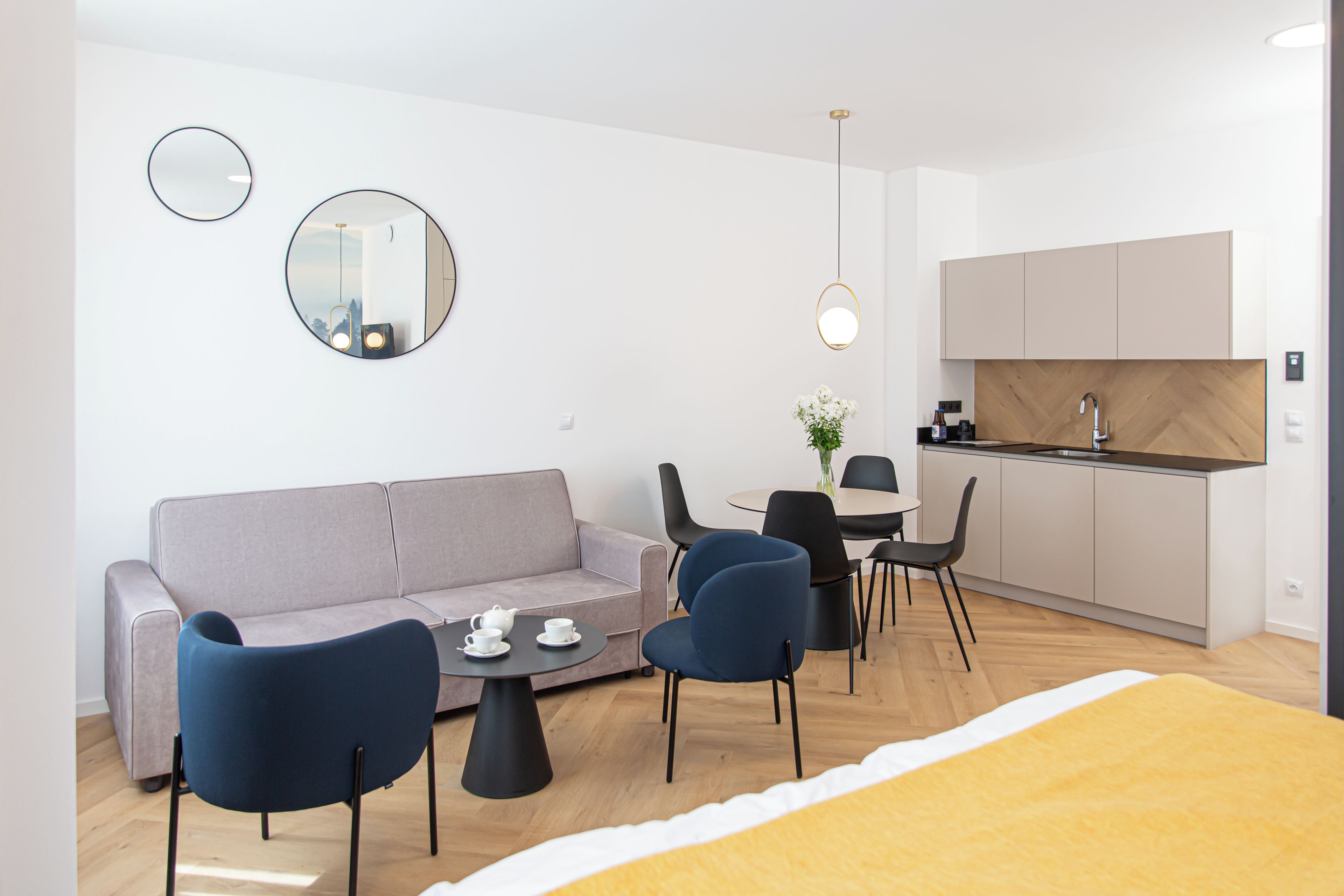 Quarto de hotel moderno com sofá, cadeiras, uma pequena mesa e cozinha compacta.