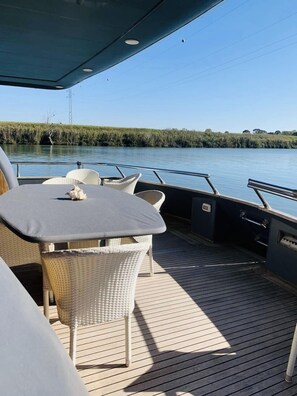 Convés de barco com mesa e cadeiras, corrimão e vista para um corpo d'água calmo com vegetação na margem.