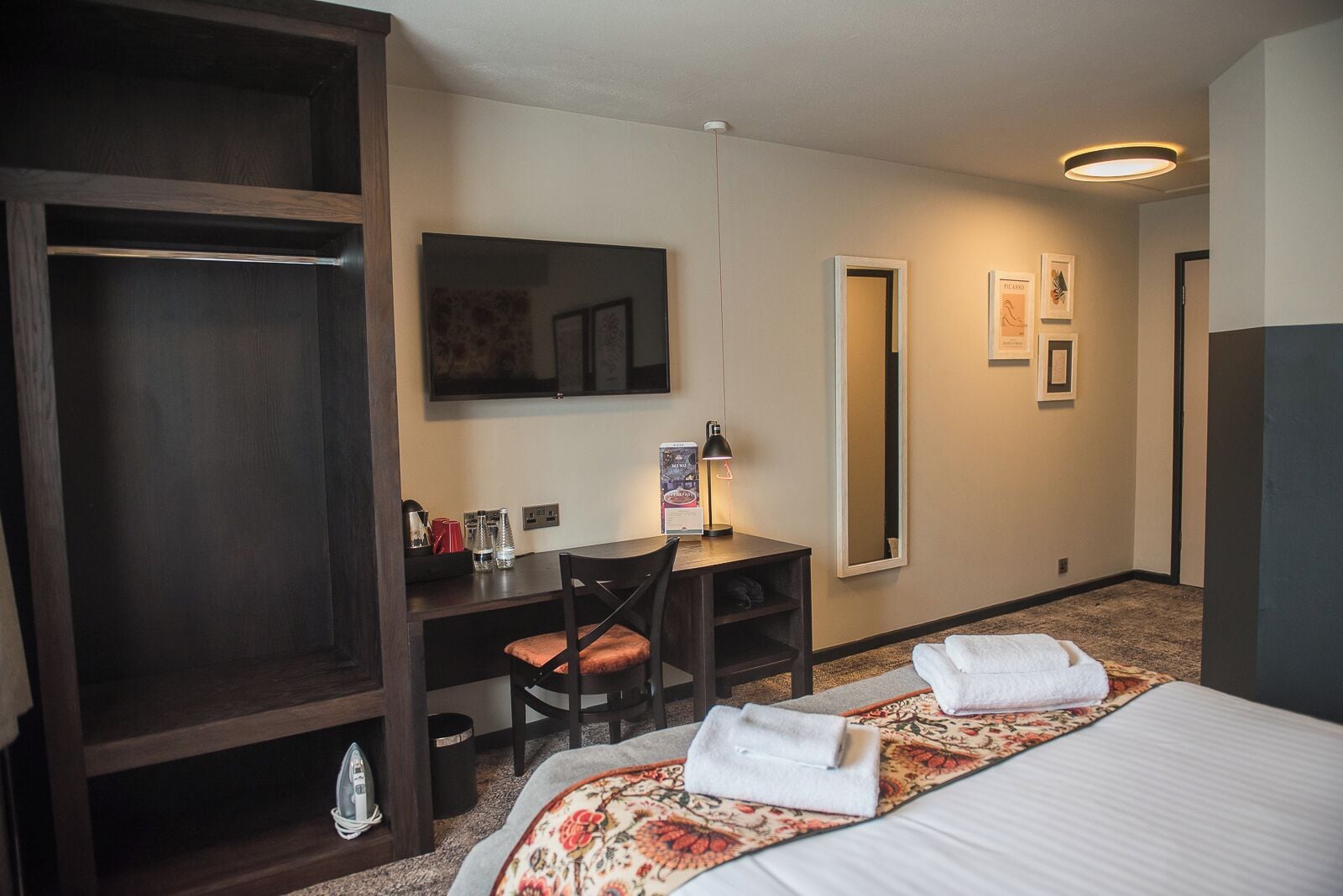Quarto de hotel com uma cama, uma escrivaninha com cadeira, um guarda-roupa, um espelho e um quadro na parede.