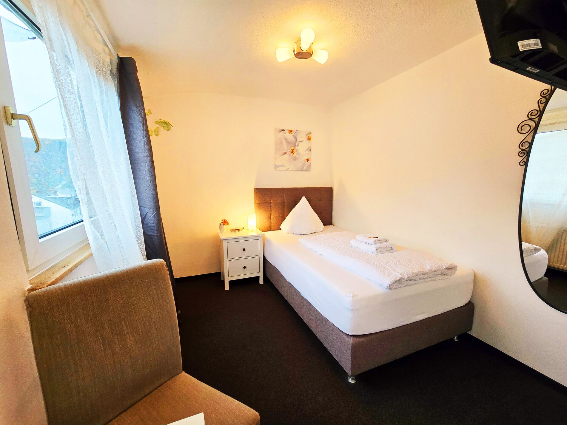 Quarto de hotel com cama, mesa de cabeceira, cadeira, janela com cortinas, TV fixada na parede e luminária no teto.