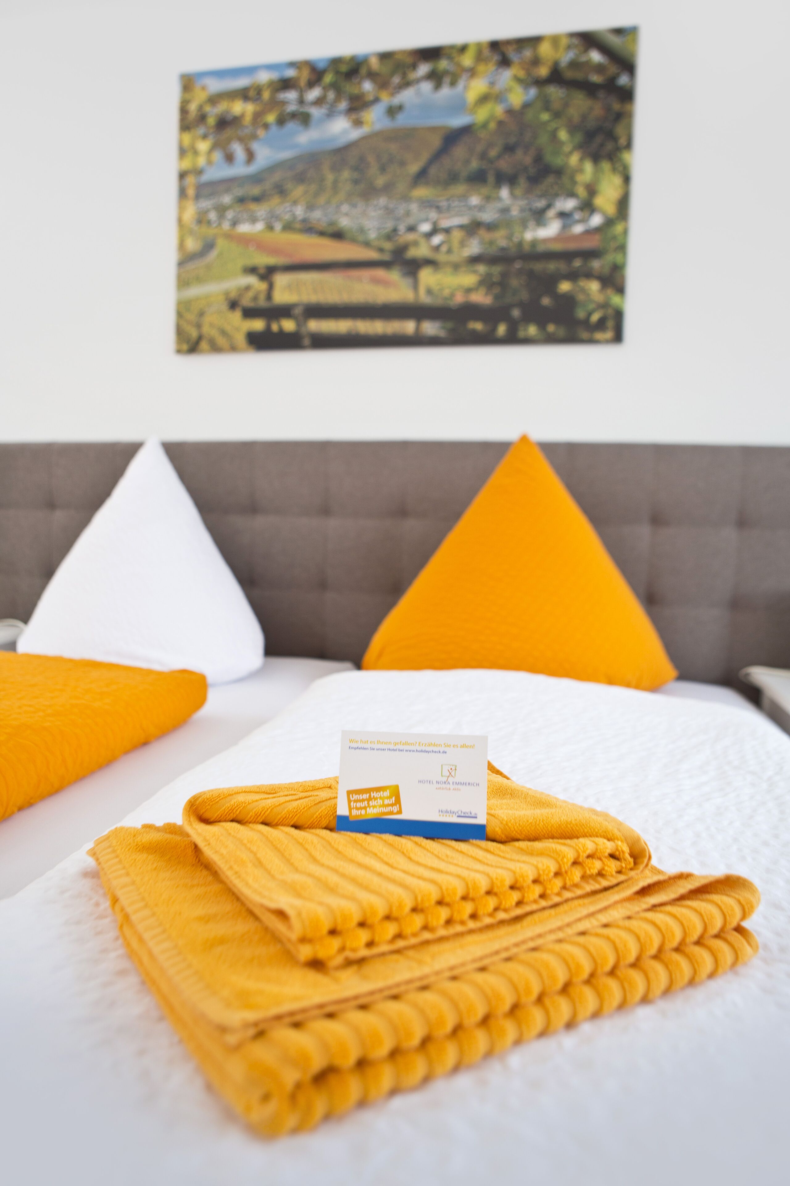 Cama bem arrumada com toalha amarela dobrada e um cartão de hotel, cabeceira cinza estofada e uma pintura de paisagem na parede.