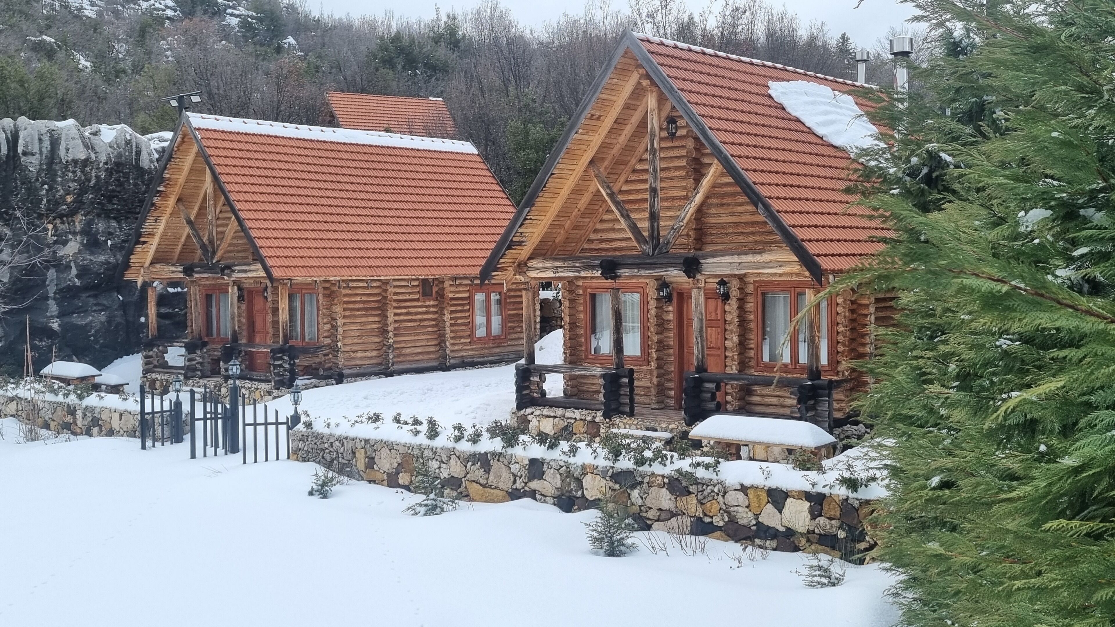 Uma cabana de troncos com alicerce de pedra e telhado de telhas vermelhas, rodeada por neve e árvores.