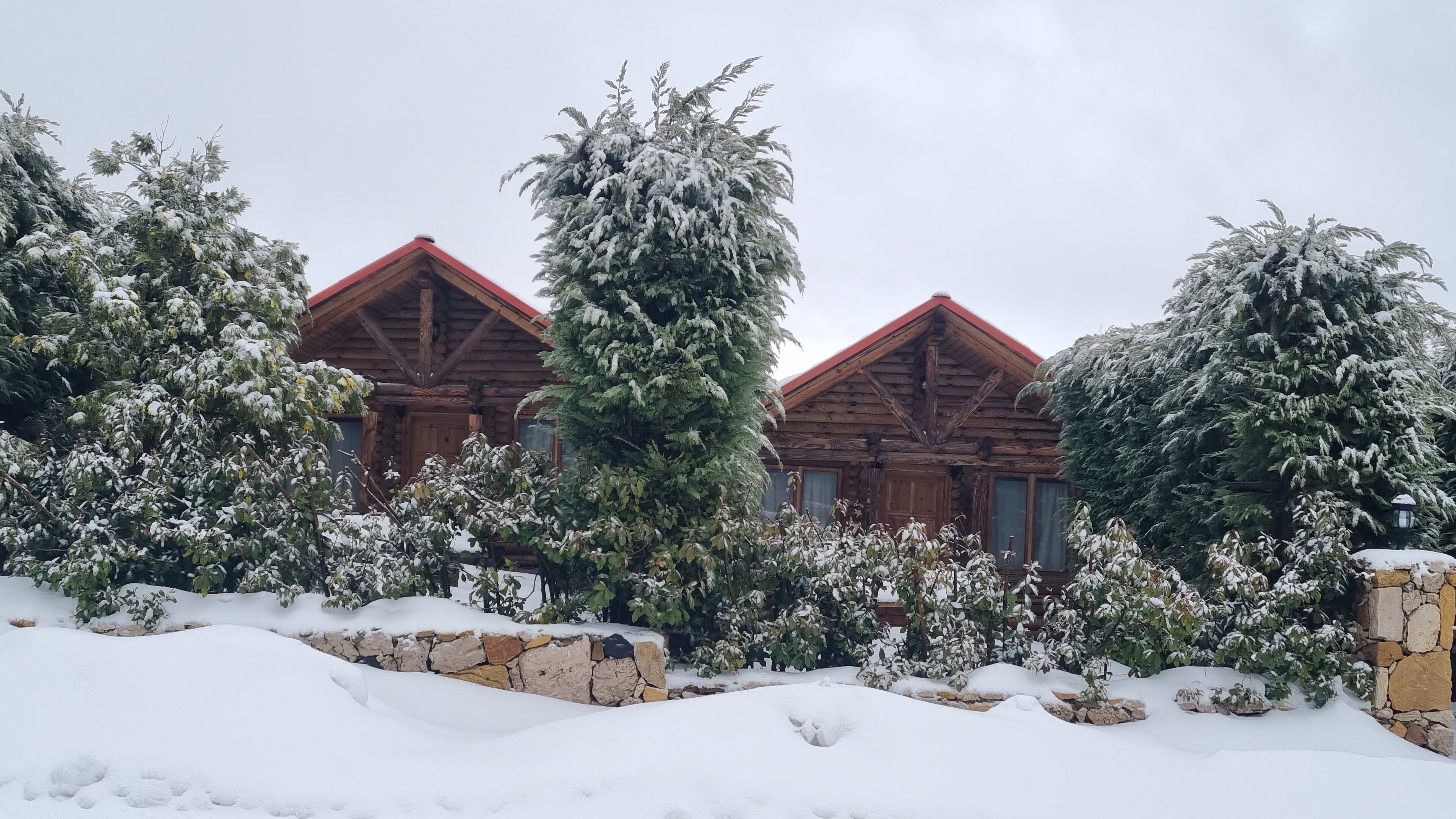 Uma cabana de tronco com telhado vermelho, rodeada por árvores cobertas de neve e uma parede de pedra.