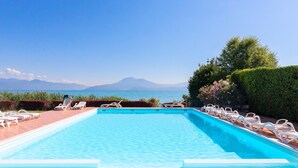 Piscina retangular com água azul cristalina, rodeada por espreguiçadeiras brancas e vista para montanhas e mar.