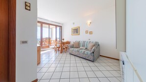 Uma sala de estar ampla com sofá, mesa de jantar e cadeiras. Há uma porta de vidro deslizante que dá para uma varanda.