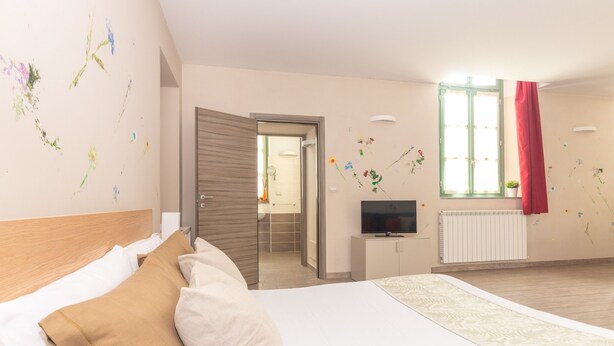 Quarto de hotel com cama, televisão, janela com cortinas, radiador e porta para um banheiro.