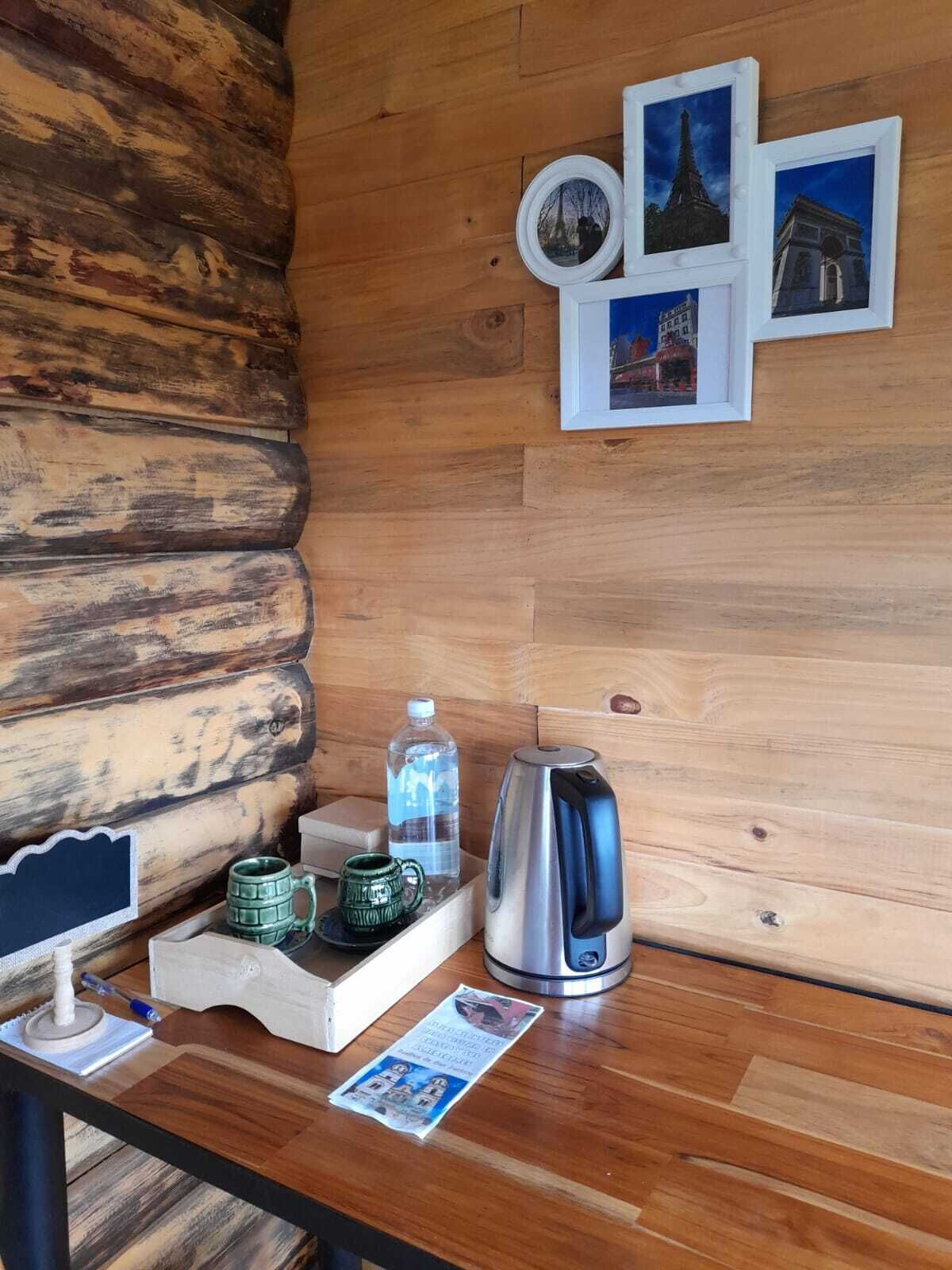 Interior de cabana de madeira com parede de madeira, mesa de madeira com chaleira, canecas e garrafa de água. Quadros emoldurados na parede.