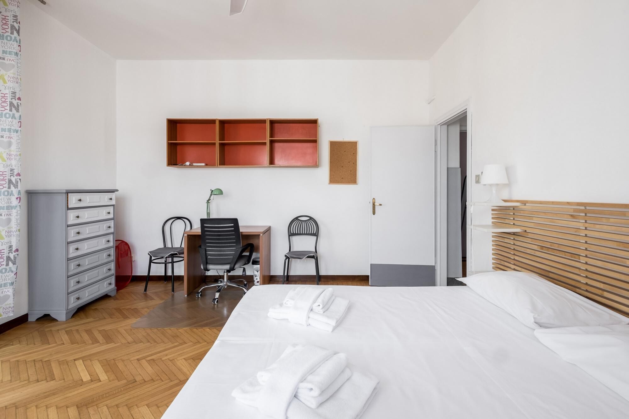 Quarto de hotel com cama, escrivaninha, duas cadeiras, guarda-roupa e prateleira na parede.