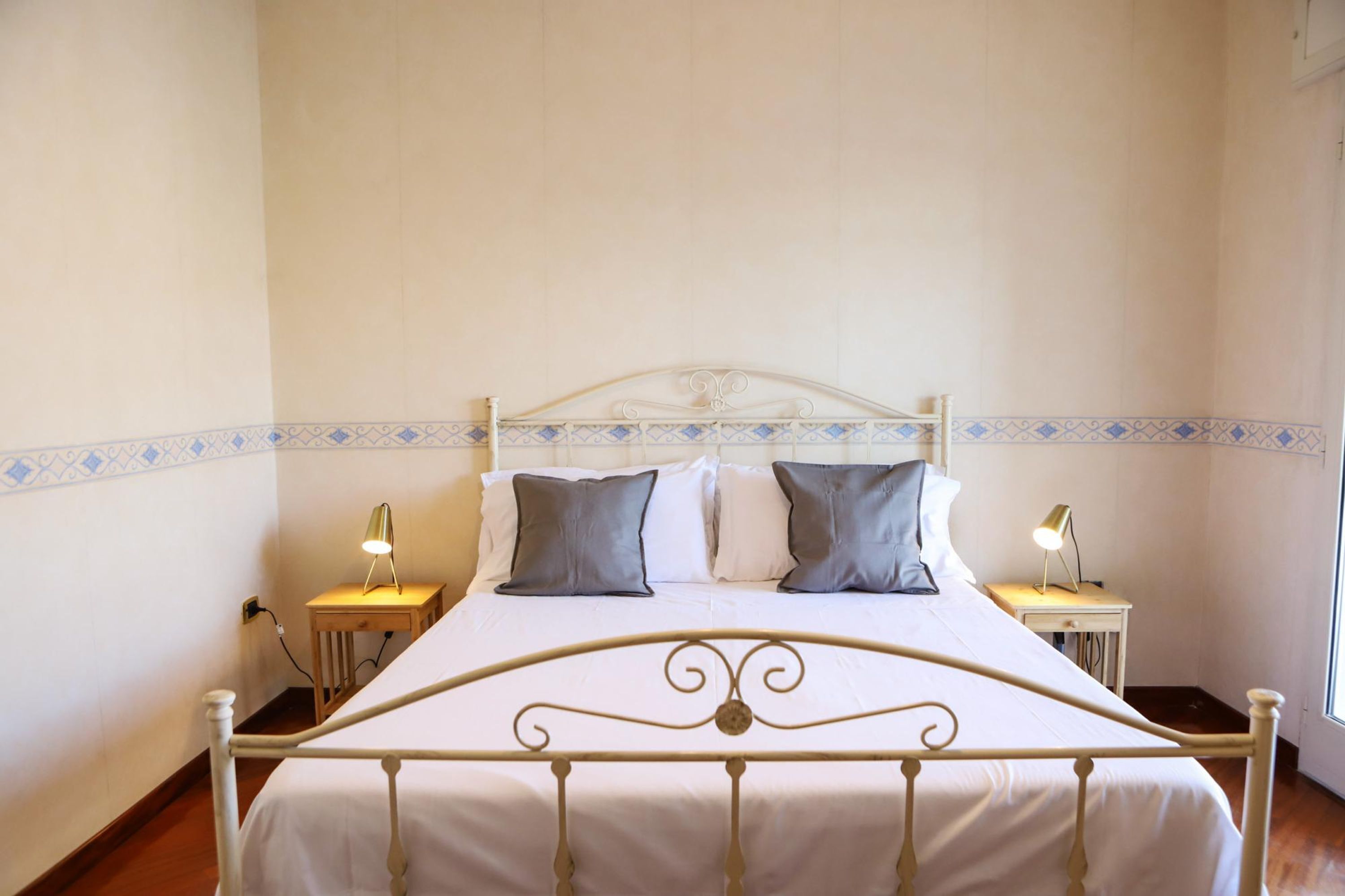 Cama bem arrumada com estrutura de metal branca e cabeceira decorativa, duas mesas de cabeceira com abajures e um padrão decorativo na parede.
