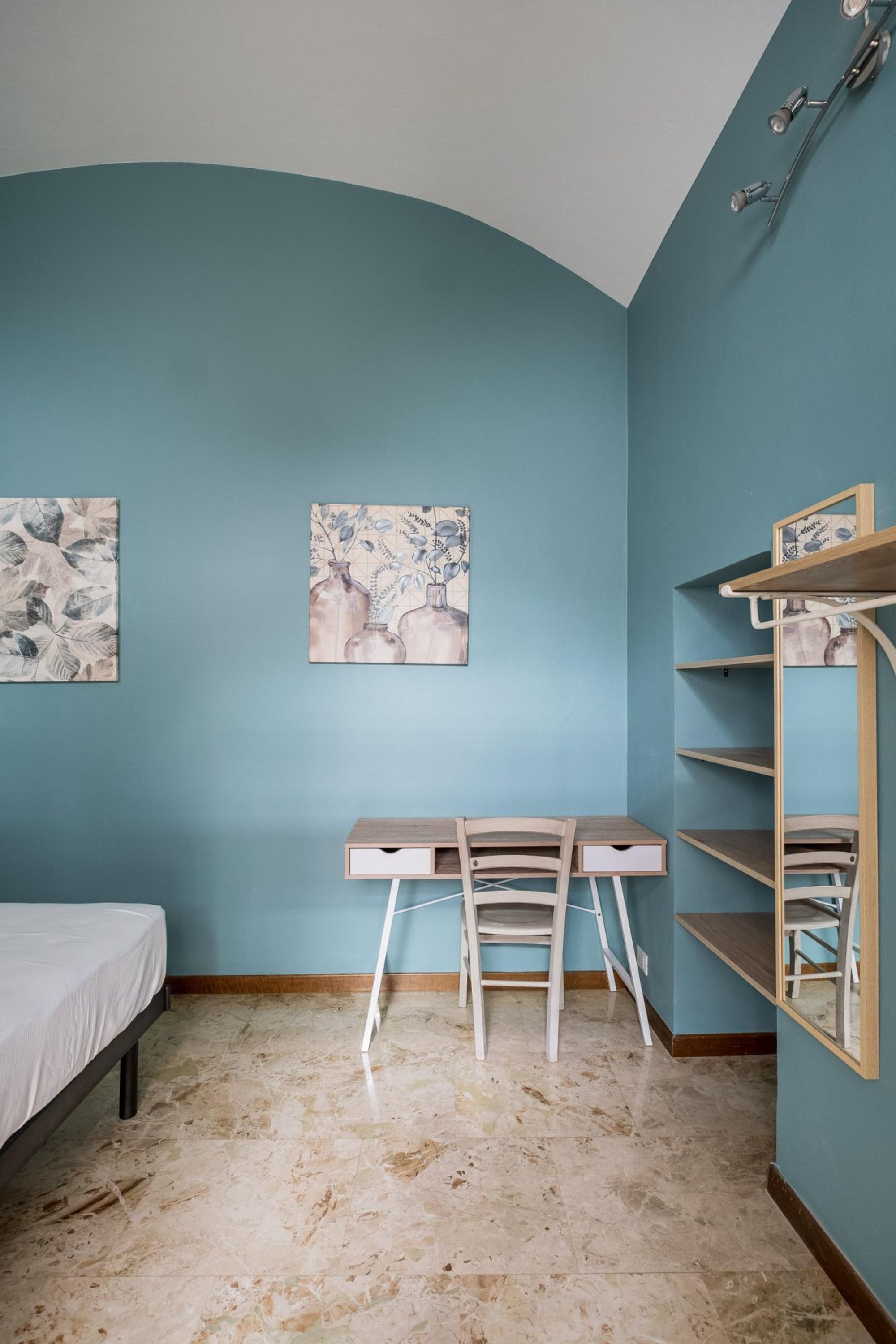 Quarto com cama, escrivaninha com cadeiras, estante e duas pinturas penduradas na parede.