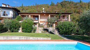 Uma casa de dois andares com piscina, rodeada por vegetação e colinas.