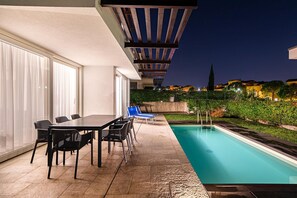 Uma área de piscina externa com mesa e cadeiras, uma pérgula e vista para a cidade à noite.