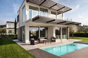 Uma casa moderna com dois andares, varanda de vidro, piscina e móveis de exterior.