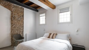 Quarto com cama, cadeira, parede de pedra e duas janelas.