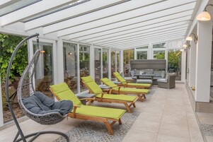 Um quarto iluminado pelo sol com teto de vidro, poltronas de madeira e cor verde, uma cadeira suspensa e um sofá cinzento.
