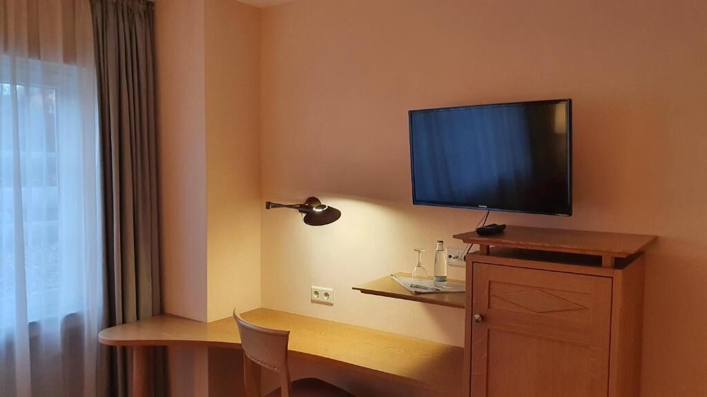 Quarto de hotel com uma mesa, uma cadeira, uma TV fixada na parede e um armário.