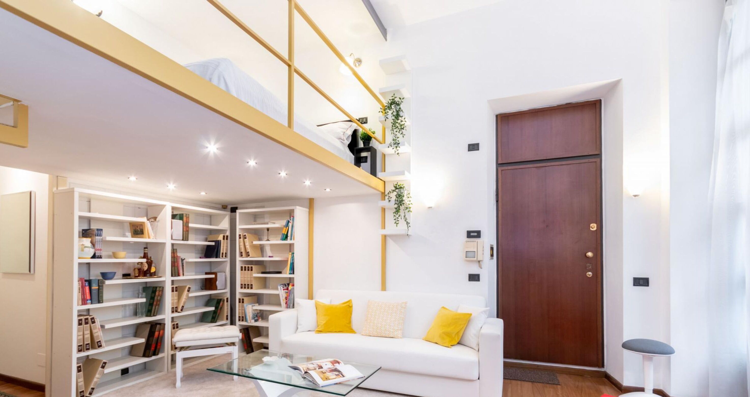 Uma sala de estar moderna com um sofá branco, almofadas amarelas, uma mesa de centro de vidro e uma estante cheia de livros e objetos decorativos.
