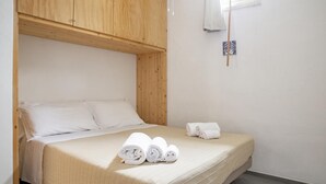 Uma cama de solteiro com um cobertor bege e duas toalhas brancas enroladas. Há uma cabeceira de madeira e uma pequena janela com cortina branca.