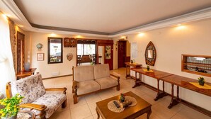 Uma sala de estar ampla com sofá, poltrona e mesa de centro. Há uma mesa de madeira com um espelho e uma janela com vista.