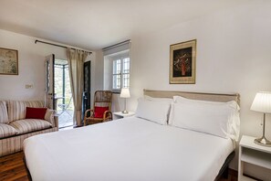 Quarto de hotel com uma cama grande, um sofá, uma poltrona, uma mesinha de cabeceira e uma janela com cortinas.