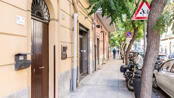 Uma rua estreita na cidade, com motocicletas e carros estacionados, um pedestre caminhando e uma placa de proibido estacionar.