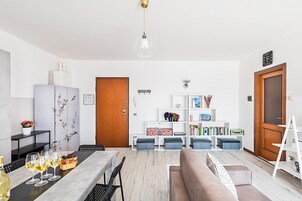 Uma sala de estar moderna com piso de madeira, um sofá cinza, uma mesa de jantar com copos de vinho e uma estante com livros e objetos decorativos.