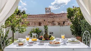 Mesa posta com itens de café da manhã, incluindo frutas, pão e bebidas, em ambiente externo com vista para um edifício histórico e céu claro.
