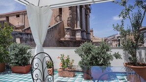 Um terraço com piso xadrez, plantas em vasos, uma cadeira de metal e vista para arquitetura histórica.